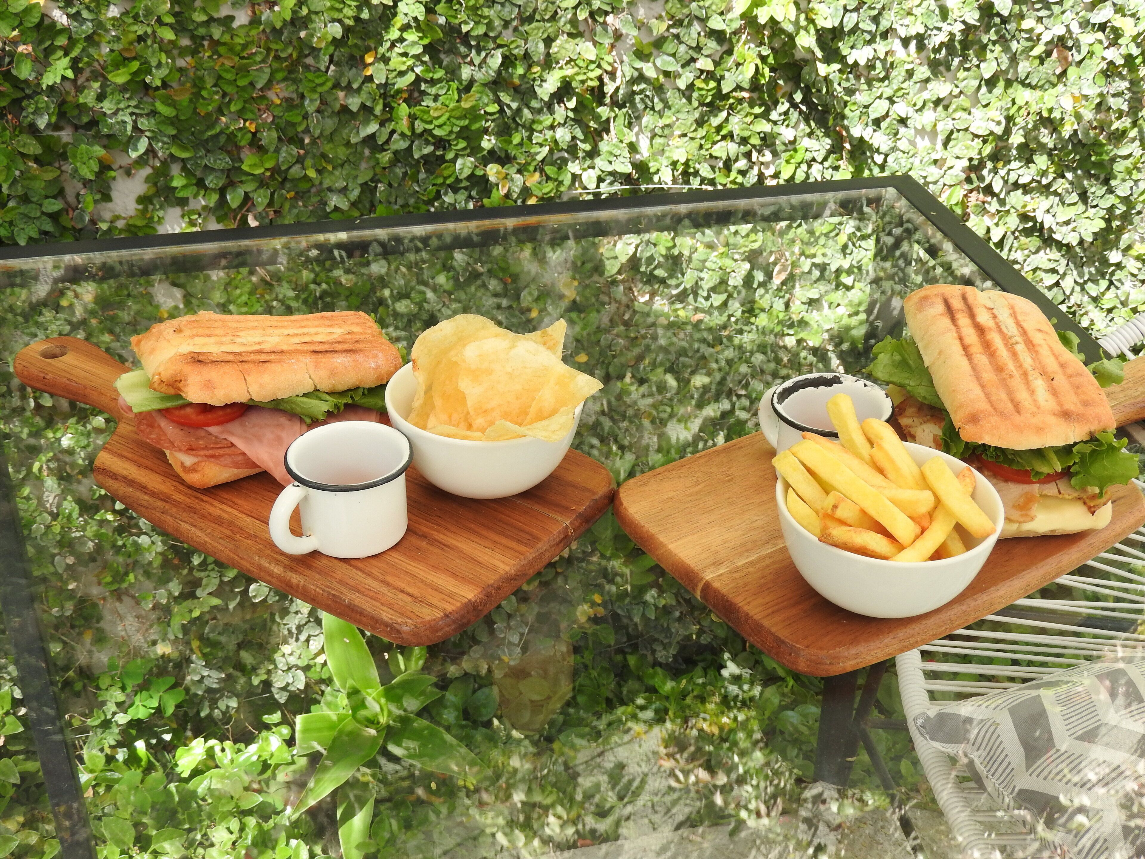 Uma mesa de vidro com duas tábuas de madeira, cada uma com um sanduíche, batatas fritas e uma chávena.
