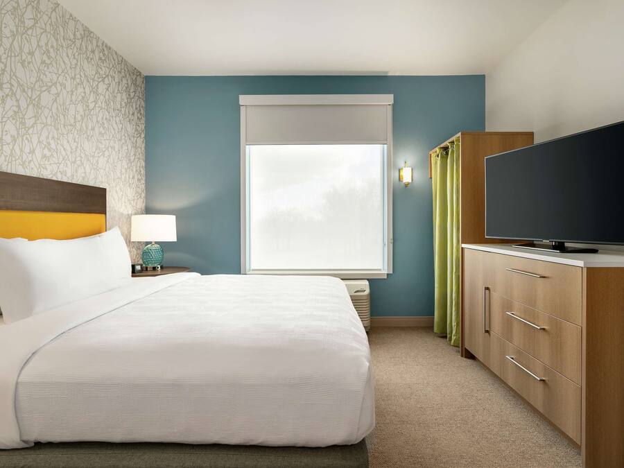 Quarto de hotel com cama, televisão, mesa de cabeceira, luminária, janela com cortinas e guarda-roupa.