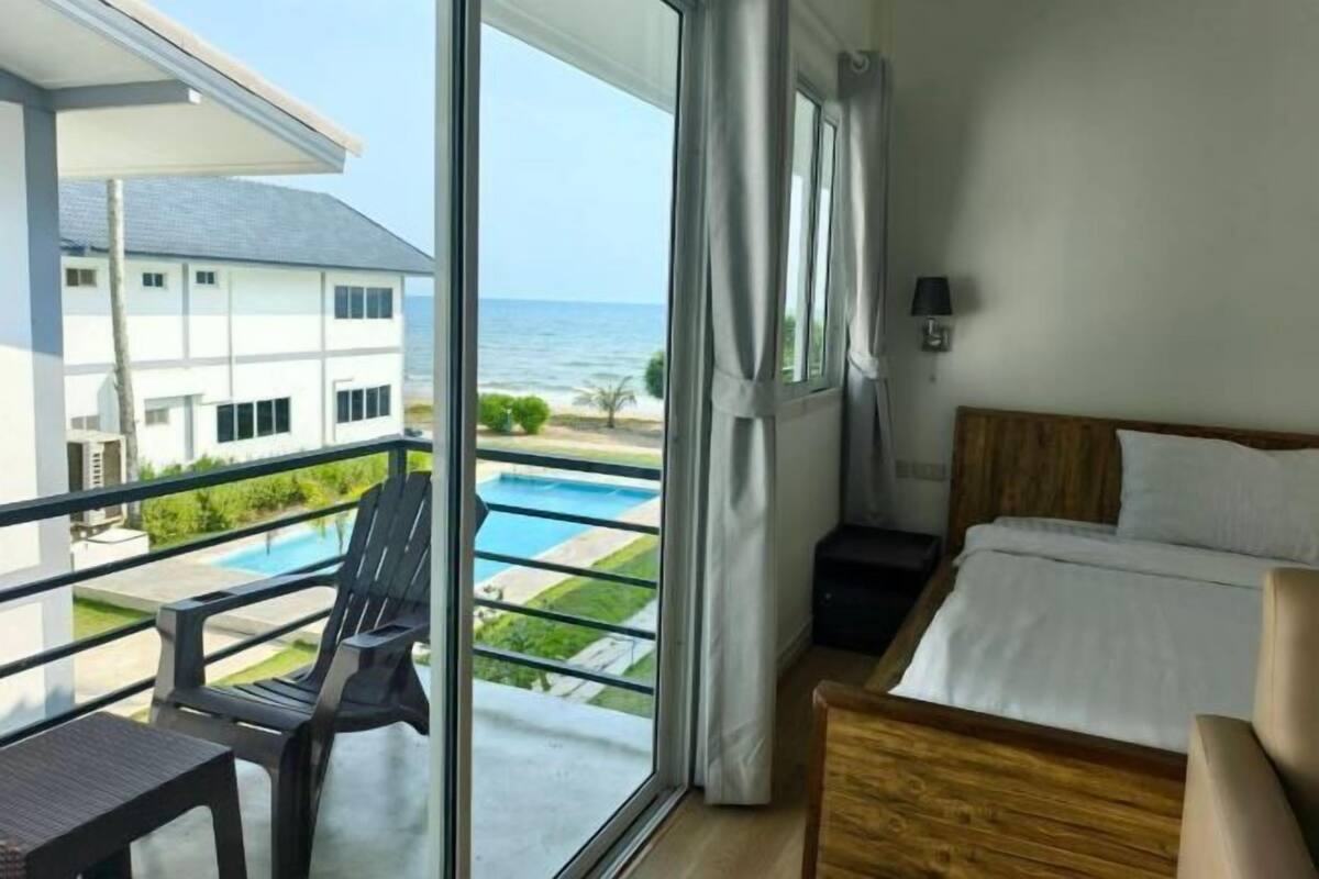 Quarto de hotel com varanda, cama, mesa de cabeceira e vista para o mar e piscina.