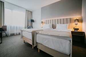 Quarto de hotel com duas camas, uma escrivaninha e uma cadeira. Há duas mesas de cabeceira com luminárias e um relógio.