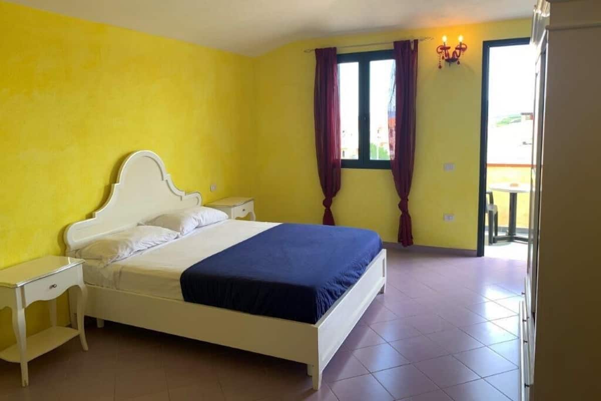Quarto com cama, mesa de cabeceira, janela com cortinas e porta para uma varanda.