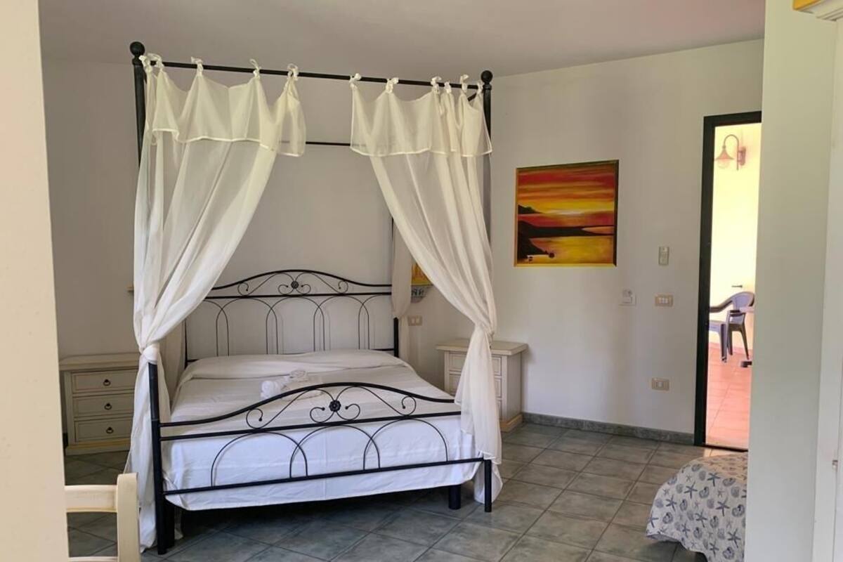 Uma cama com dossel, uma mesinha de cabeceira, uma cadeira e um quadro na parede.