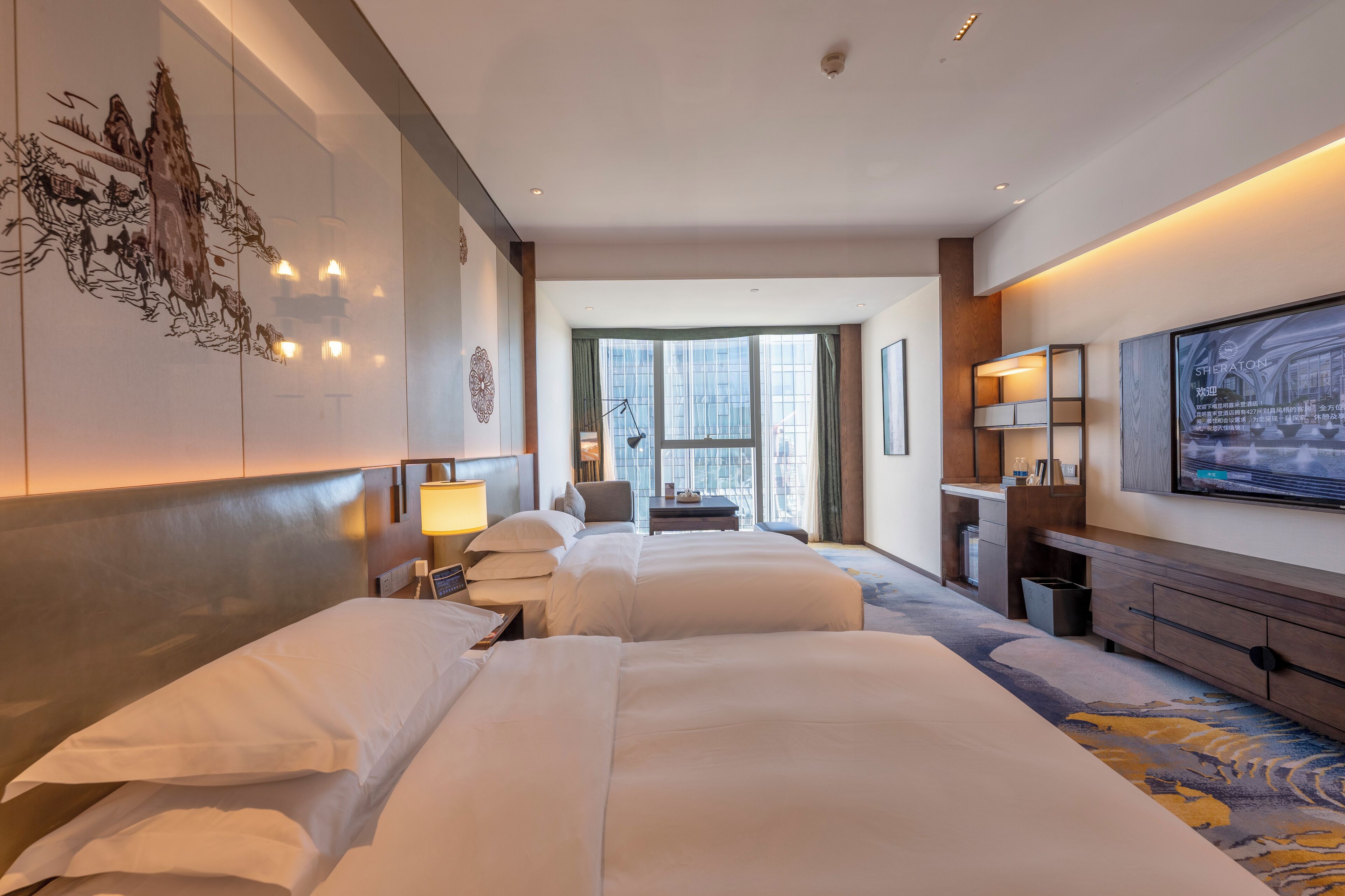 Quarto de hotel com duas camas, uma mesa, uma cadeira, televisão e uma janela grande com vista para prédios.