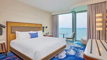 Quarto de hotel com uma cama grande, uma escrivaninha, uma cadeira, uma mesa pequena e vista para o mar através de uma janela ampla.