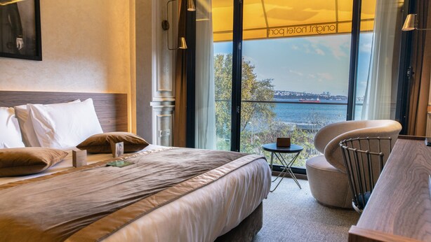 Quarto de hotel com uma cama grande, uma mesa redonda pequena, uma cadeira e vista para um corpo de água através de grandes janelas.