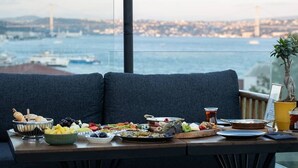 Mesa de jantar com diversos alimentos e um vaso de planta em varanda com vista para a cidade e o mar.