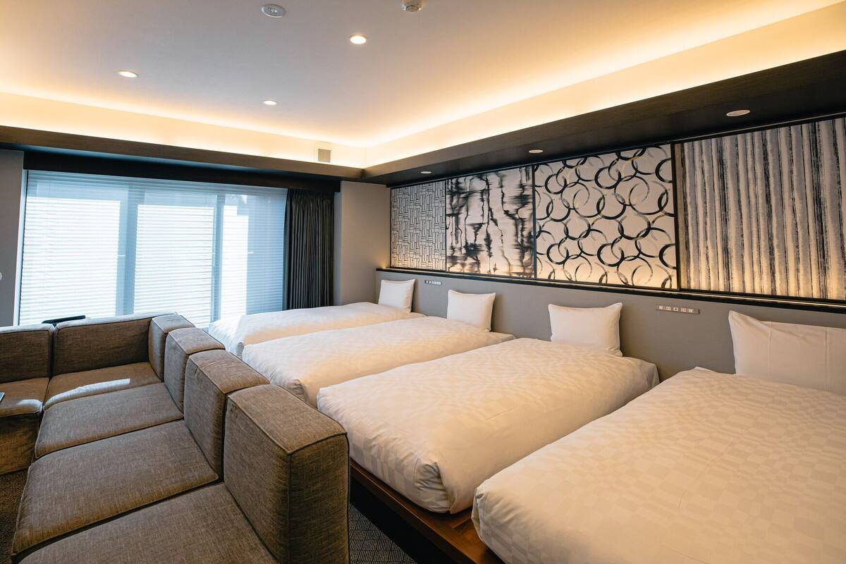 Quarto de hotel com três camas, um sofá e uma janela grande.