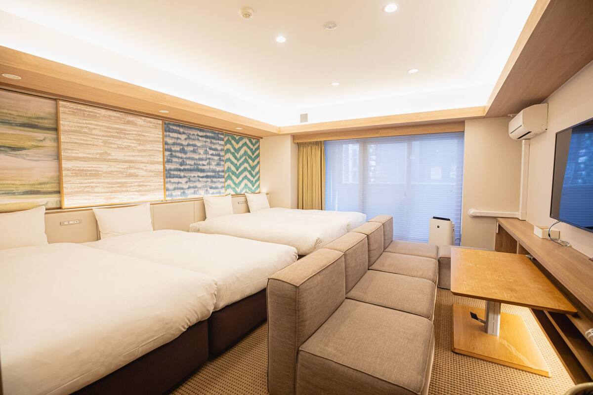 Quarto de hotel com duas camas, um sofá, uma televisão e uma janela grande.