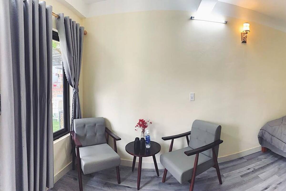 Quarto de hotel com duas camas de solteiro, uma mesinha com cadeiras, uma janela com cortinas e um ar-condicionado fixado na parede.