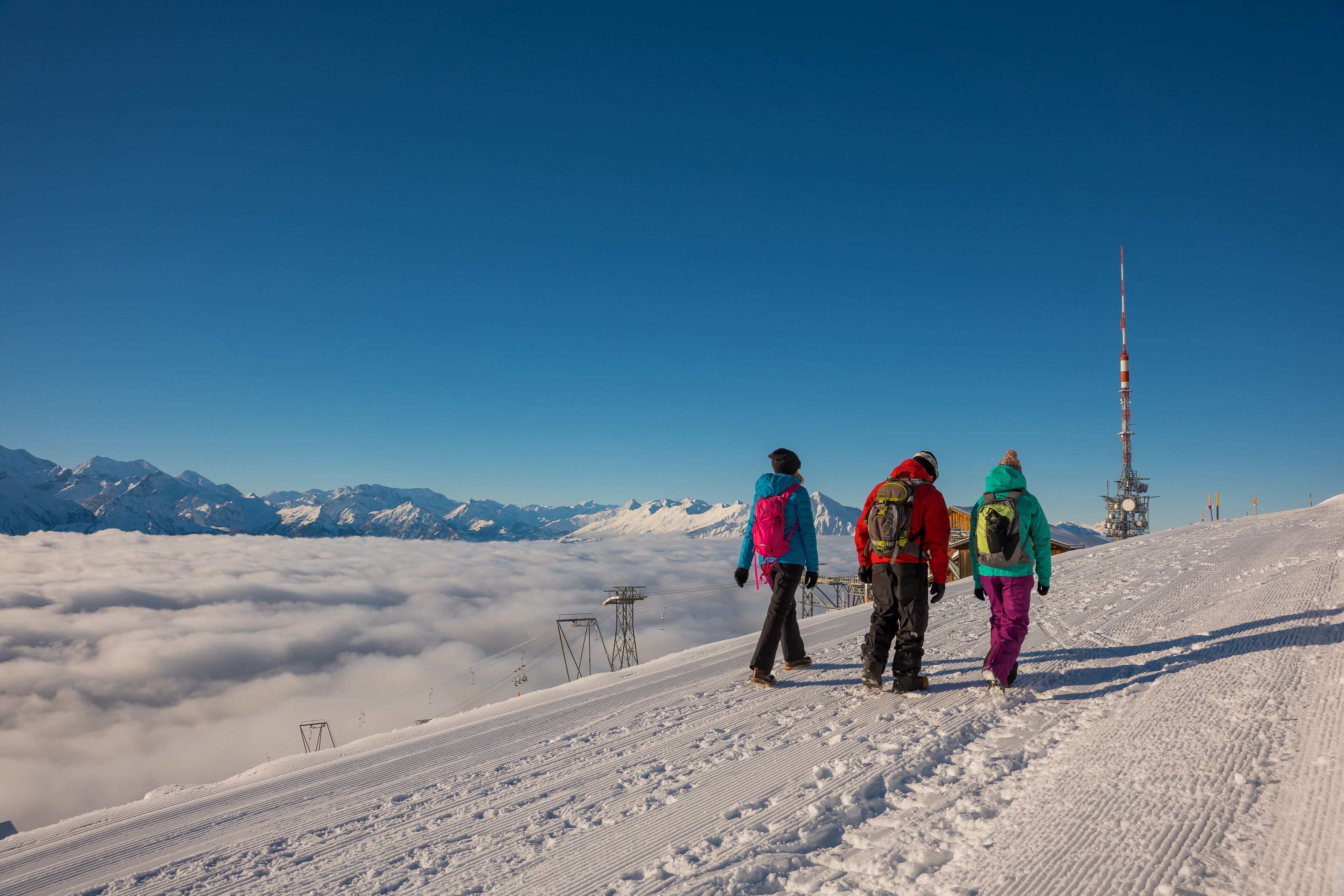 Encosta nevada de montanha com três pessoas caminhando, um teleférico ao fundo e uma torre de comunicação.