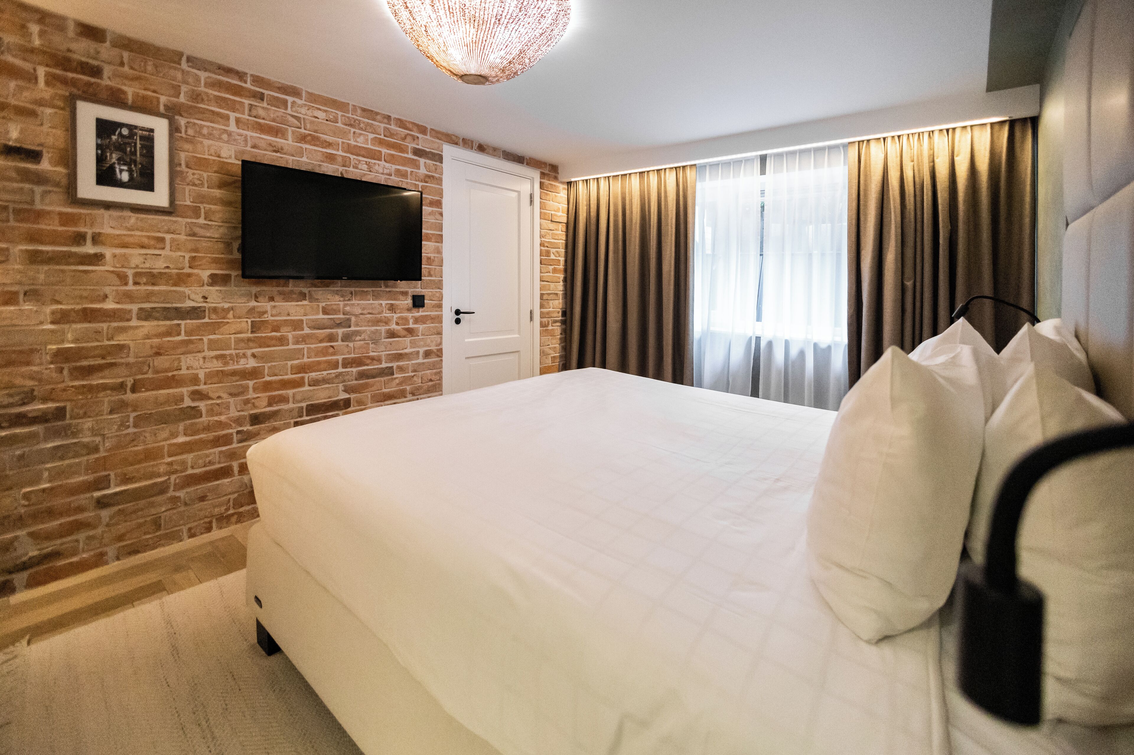 Quarto de hotel com parede de tijolos, televisão de tela plana acima de uma porta, cama grande com roupa de cama branca e uma janela com cortinas.