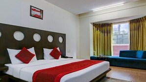 Quarto de hotel com uma cama grande, cabeceira com recortes circulares, sofá azul e uma janela com cortinas.