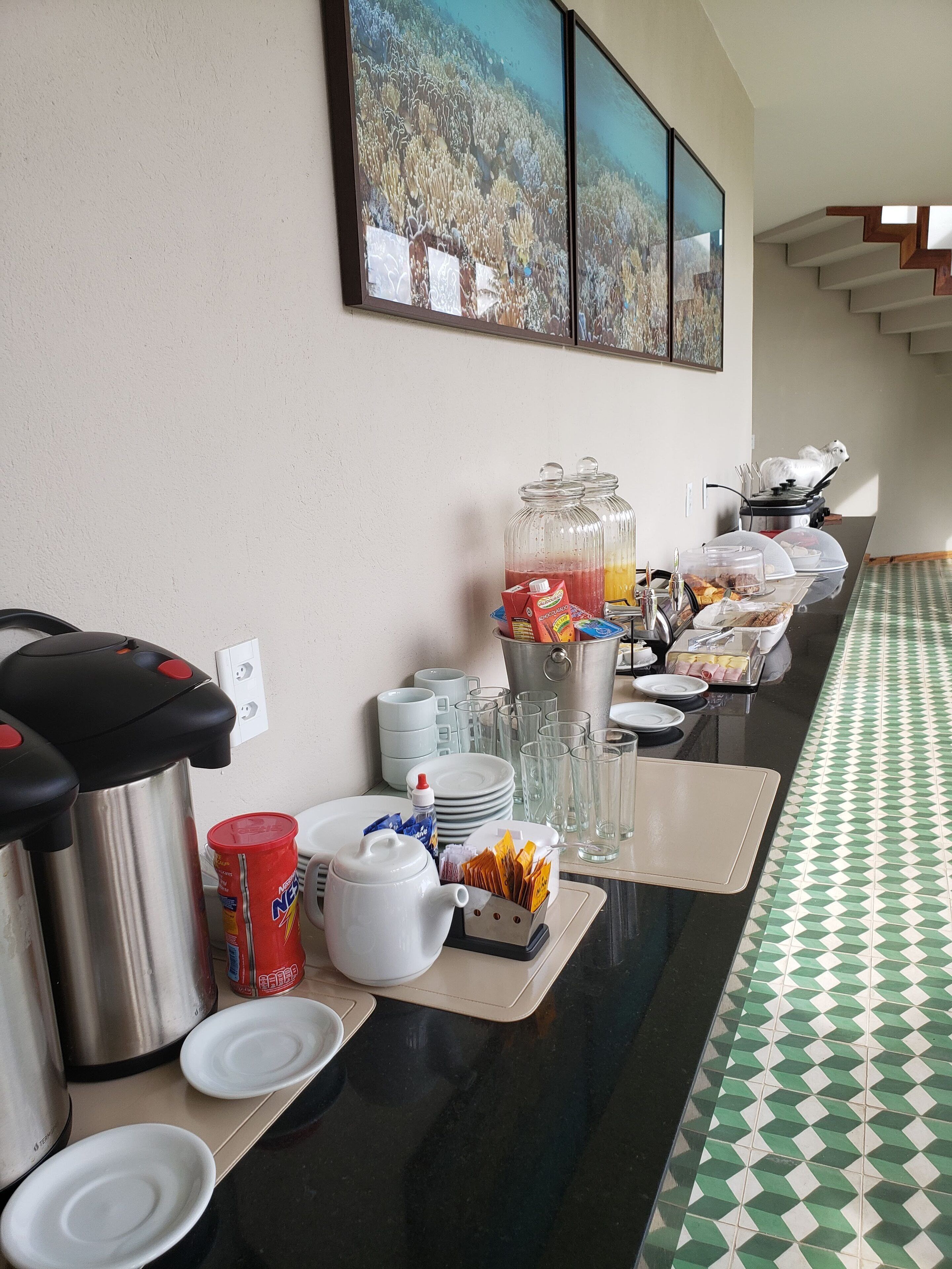 Disposição de buffet de café da manhã com diversos alimentos e bebidas, uma máquina de café e um cachorro em uma cadeira.
