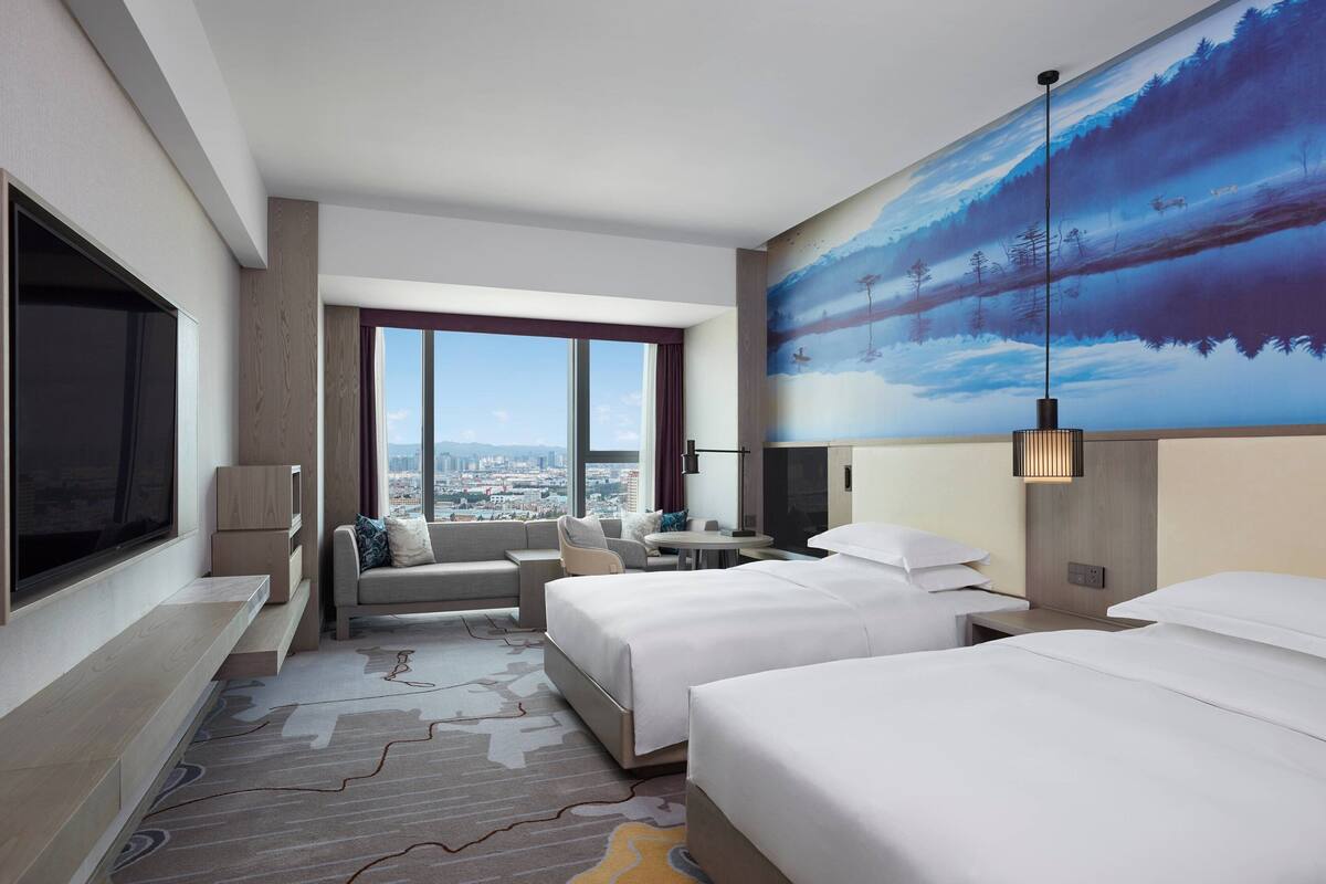 Quarto de hotel com uma janela grande, duas camas, um sofá, uma televisão e um quadro.