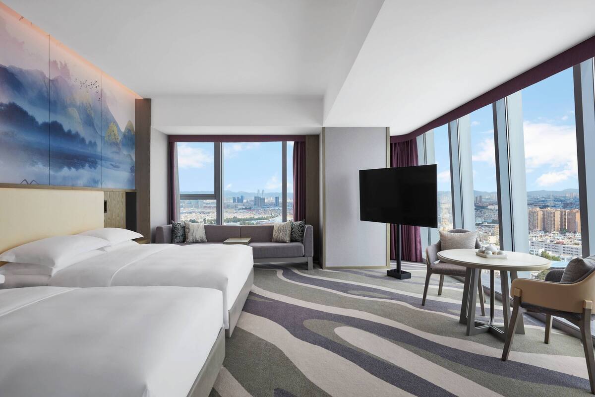 Quarto de hotel com duas camas, um sofá, televisão e vista para a cidade.
