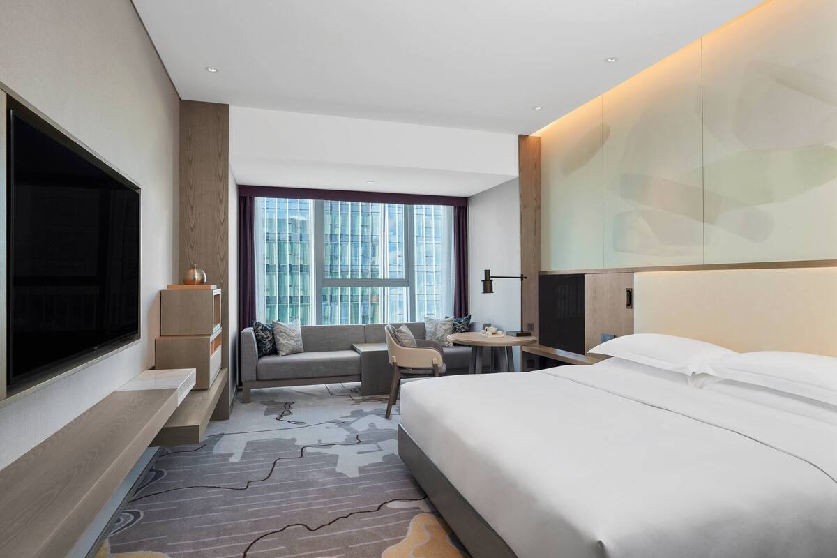 Quarto de hotel moderno com uma cama grande, área de estar com sofá e poltrona, uma mesinha e uma televisão.