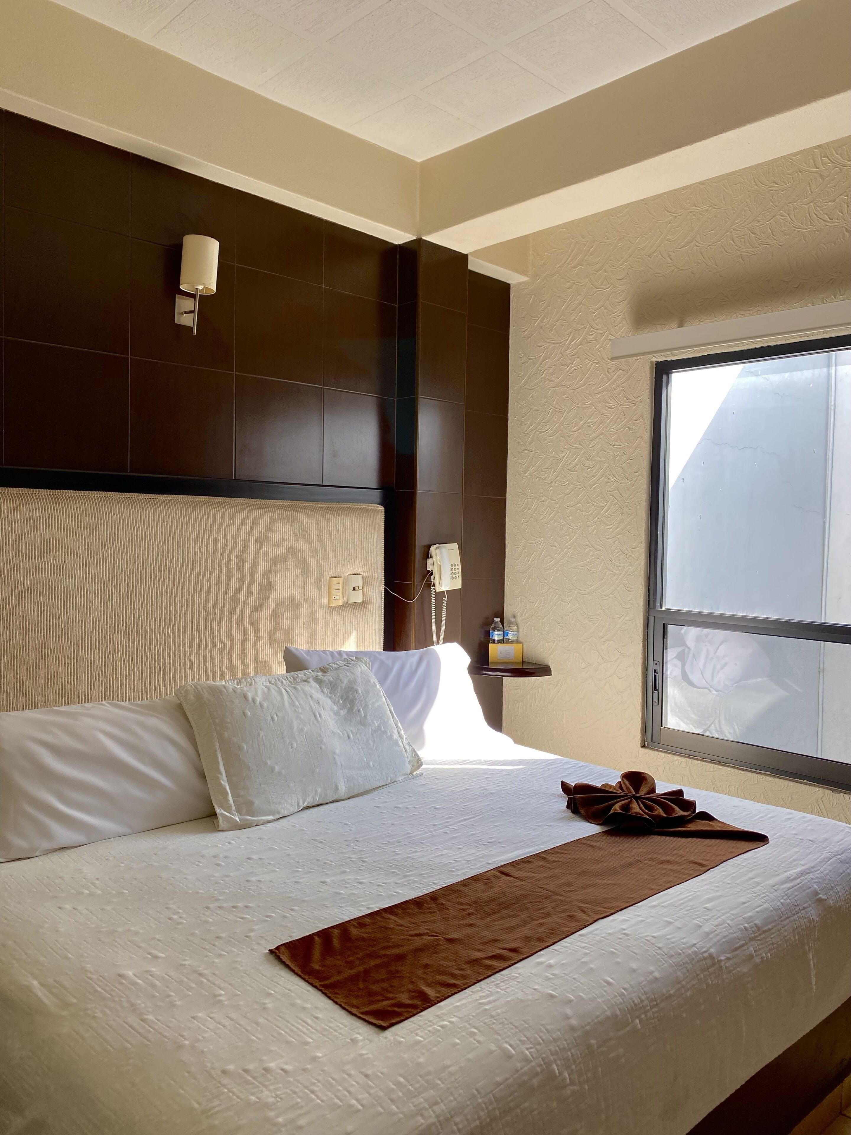Quarto de hotel com cama grande, cabeceira, mesa de cabeceira com telefone e garrafa, e janela com vista.