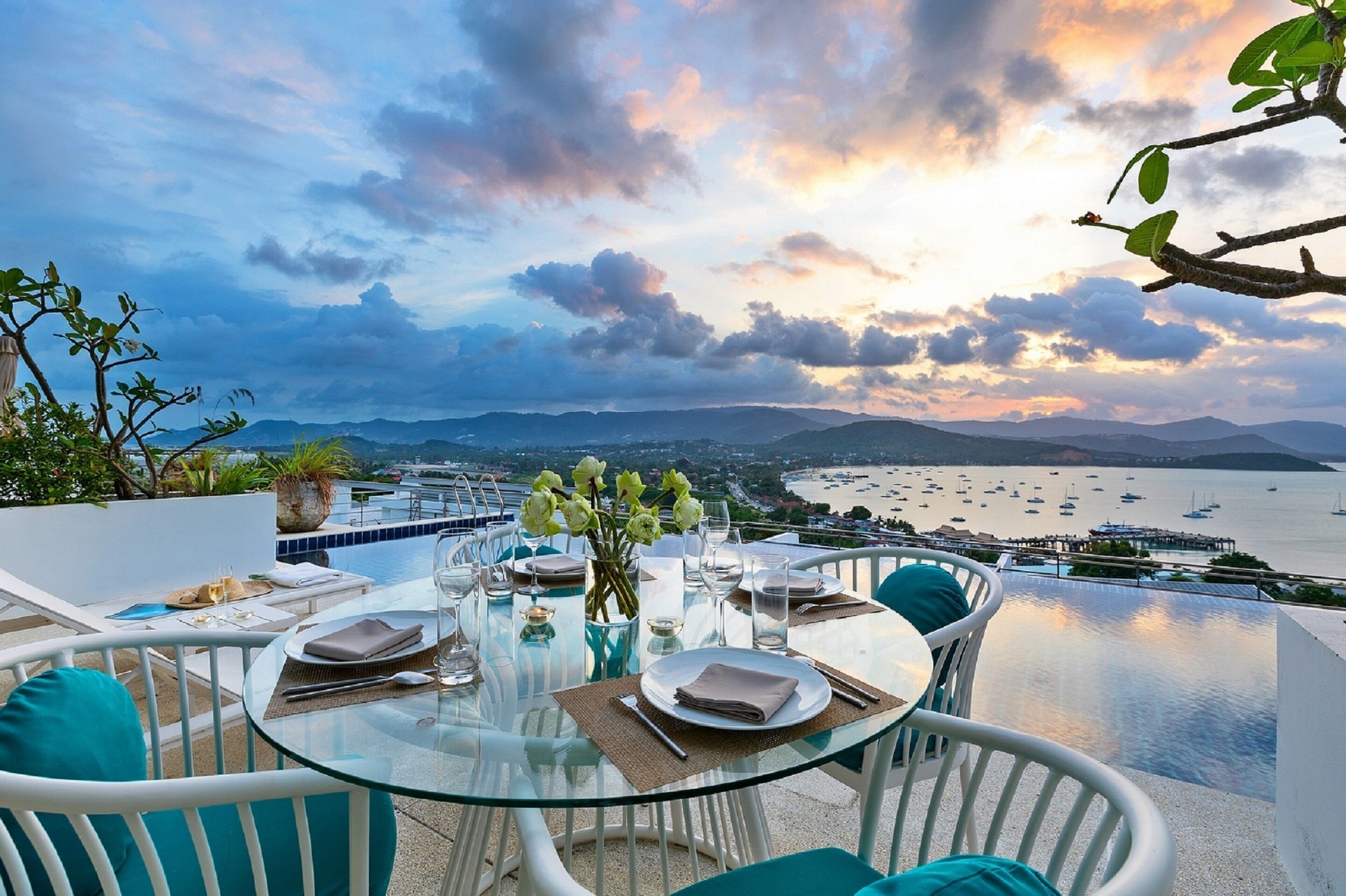 Mesa com tampo de vidro posta para refeições, com vista para o mar e barcos.