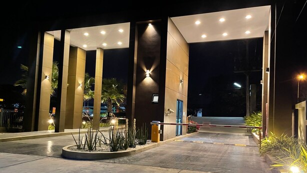 Entrada moderna de um edifício, com acesso por alameda murada, iluminada por luzes e cercada por palmeiras e plantas.