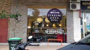 Exterior do Hotel Oyasumi, com entrada de vidro, uma placa exibindo o nome do hotel e informações de contato, e uma motocicleta estacionada em frente.
