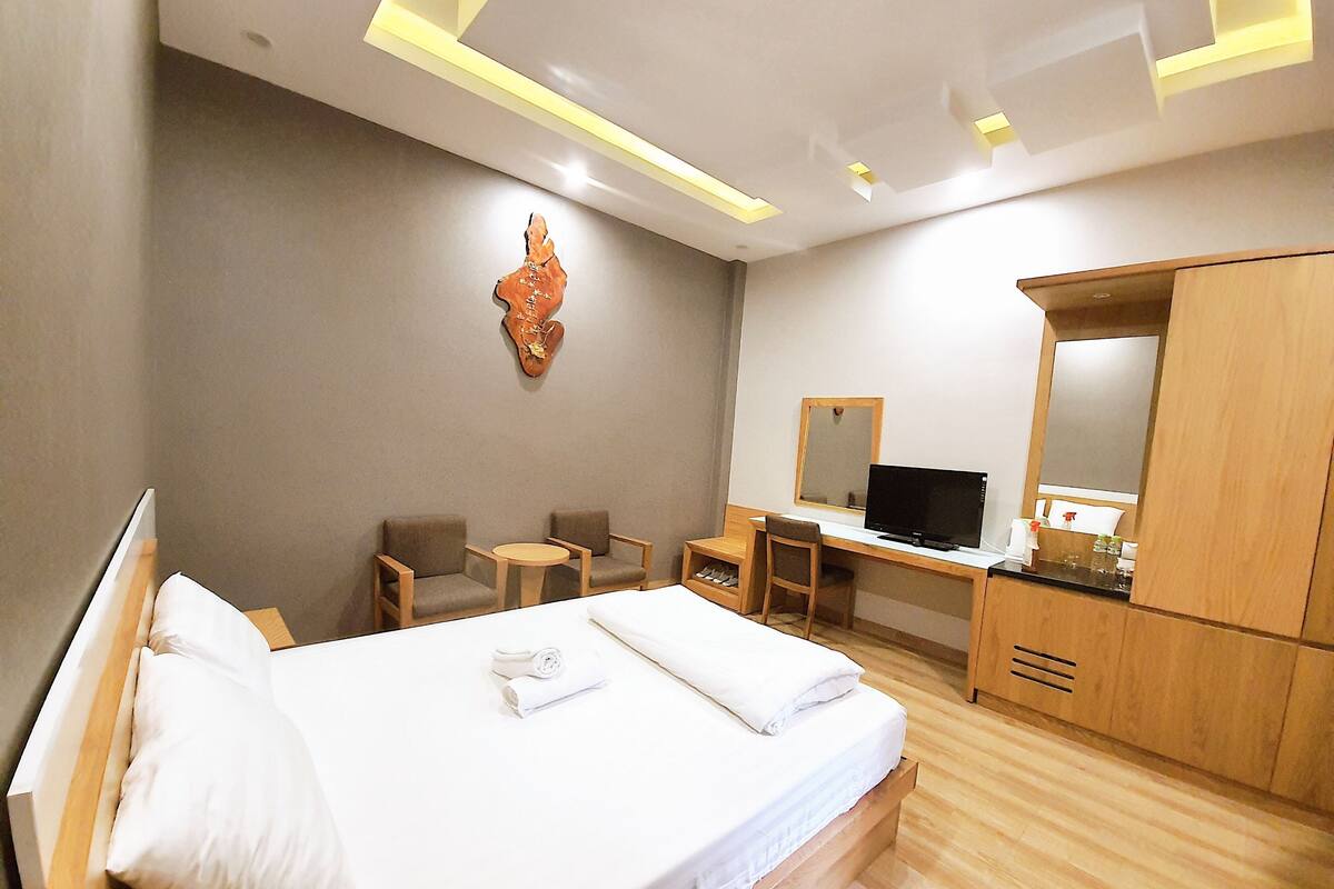 Quarto de hotel com uma cama, duas cadeiras, uma escrivaninha, televisão e um guarda-roupa.