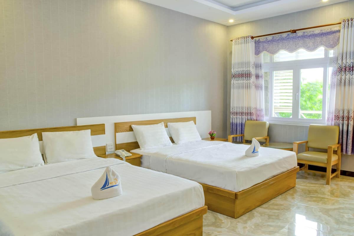 Quarto de hotel com duas camas, uma mesa pequena, uma cadeira e uma janela com cortinas.