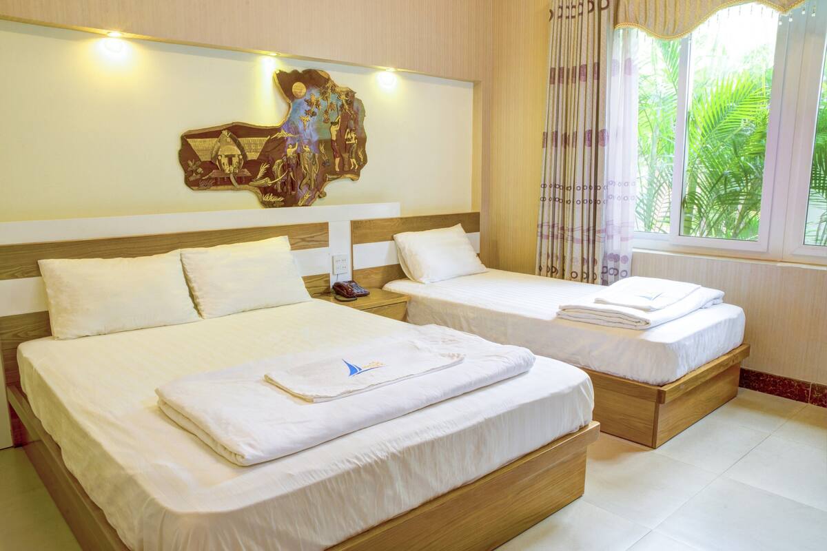 Quarto de hotel com duas camas, um objeto decorativo na parede e janela com vista para vegetação.