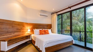 Quarto com cabeceira de madeira, cama grande com roupa de cama branca, mesa de cabeceira com abajur e vista para o verde através de grandes janelas.