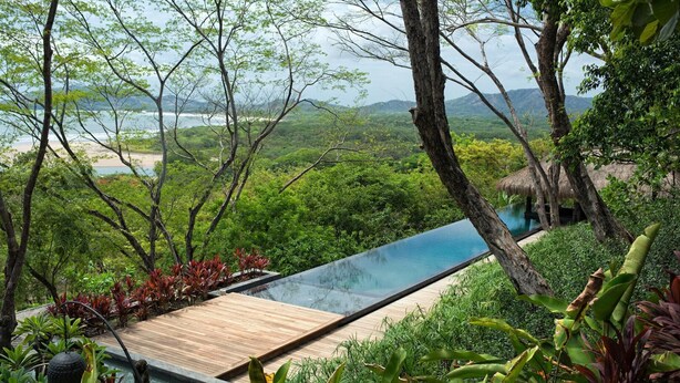 Uma piscina infinita luxuosa ao ar livre com um deck de madeira, rodeada por vegetação exuberante e com vista para uma paisagem deslumbrante.