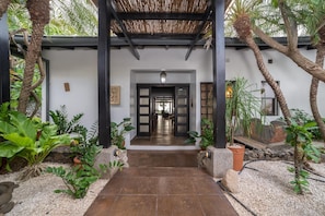 Uma entrada coberta com um caminho que leva a um edifício, cercado por plantas tropicais e um muro de pedra.