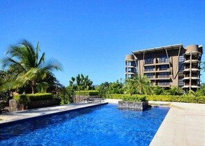 Piscina adjacente a um edifício moderno de vários andares com varandas, rodeada por palmeiras e vegetação.