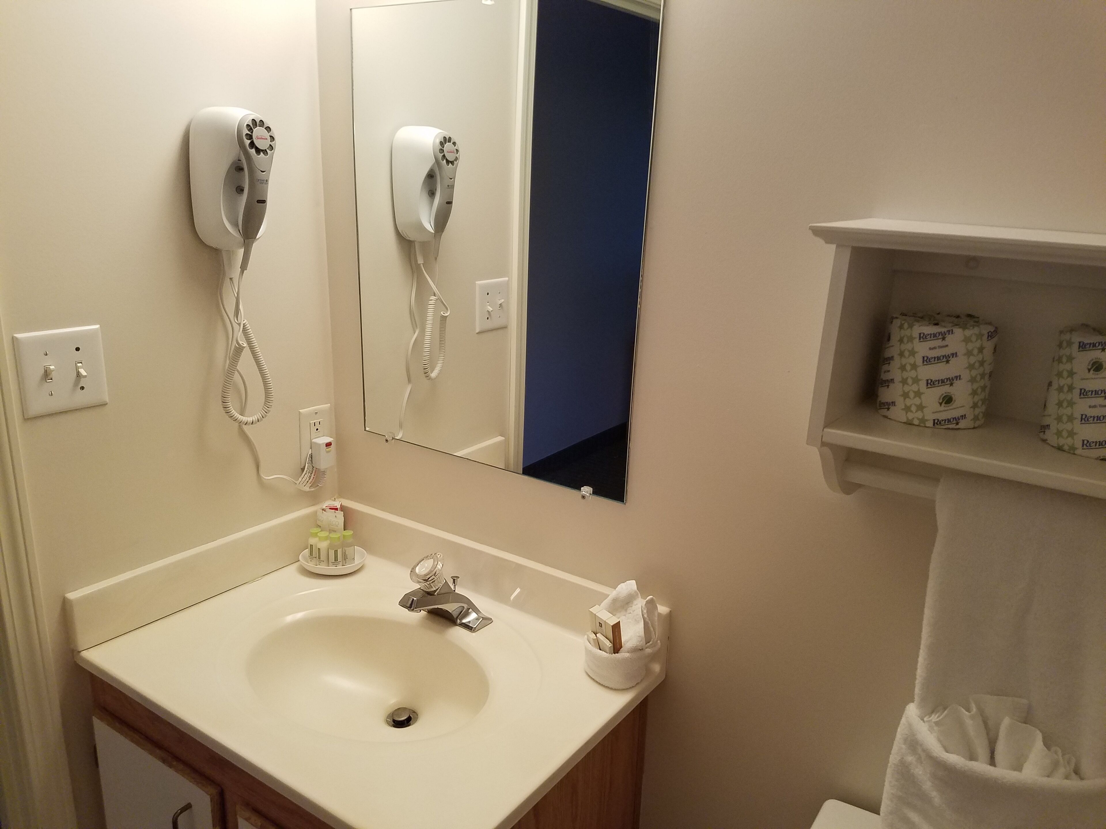 Casa de banho com lavatório, secador de cabelo, espelho e prateleira com papel higiénico.