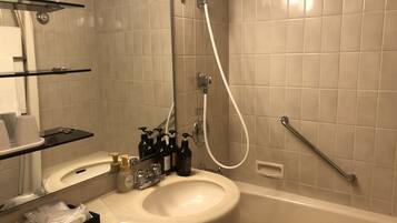 Casa de banho com lavatório branco, espelho e chuveiro. Há artigos de higiene pessoal sobre o lavatório e um porta-toalhas acima.