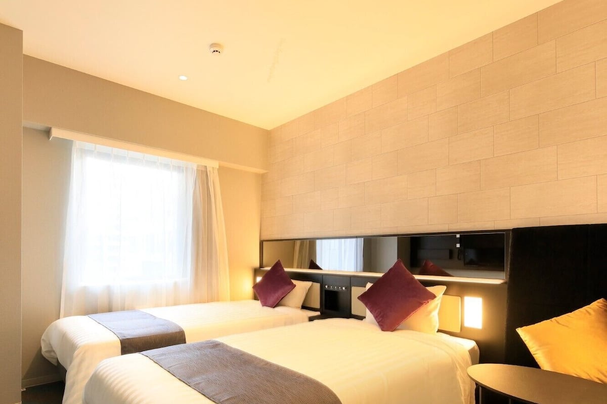 Quarto de hotel com duas camas, televisão, uma mesa pequena e janela com cortinas.
