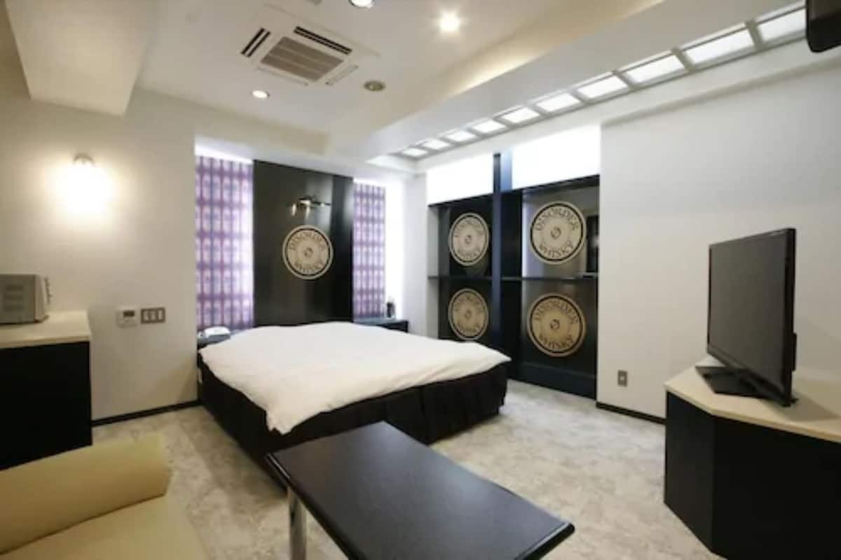 Quarto de hotel moderno com cama, escrivaninha, televisão e relógios decorativos na parede.