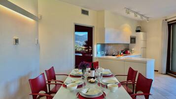 Uma sala de jantar com mesa posta para seis pessoas, cozinha ao fundo e vista para montanhas por uma porta aberta.