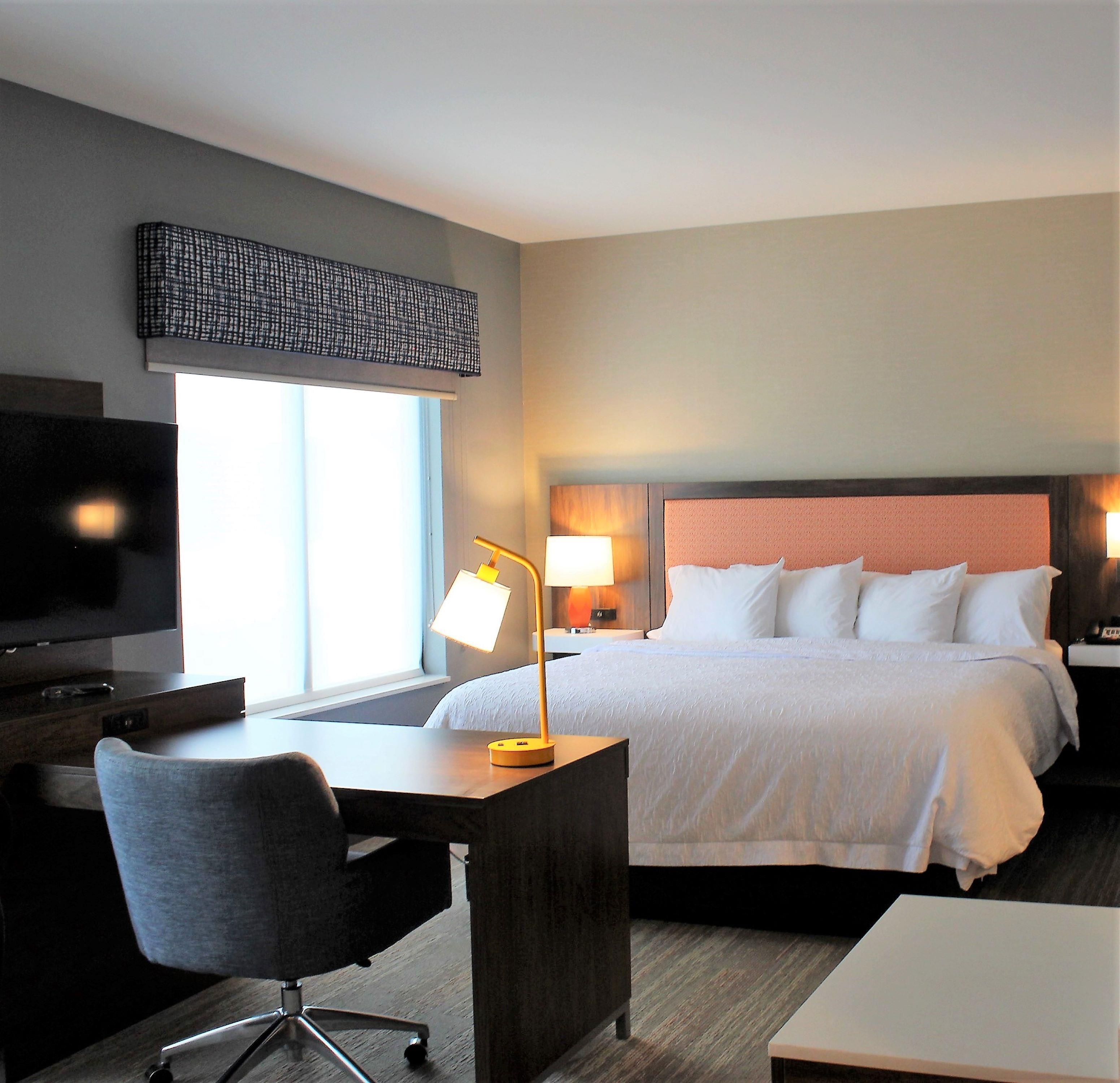 Quarto de hotel com cama, escrivaninha, cadeira, televisão e janela.