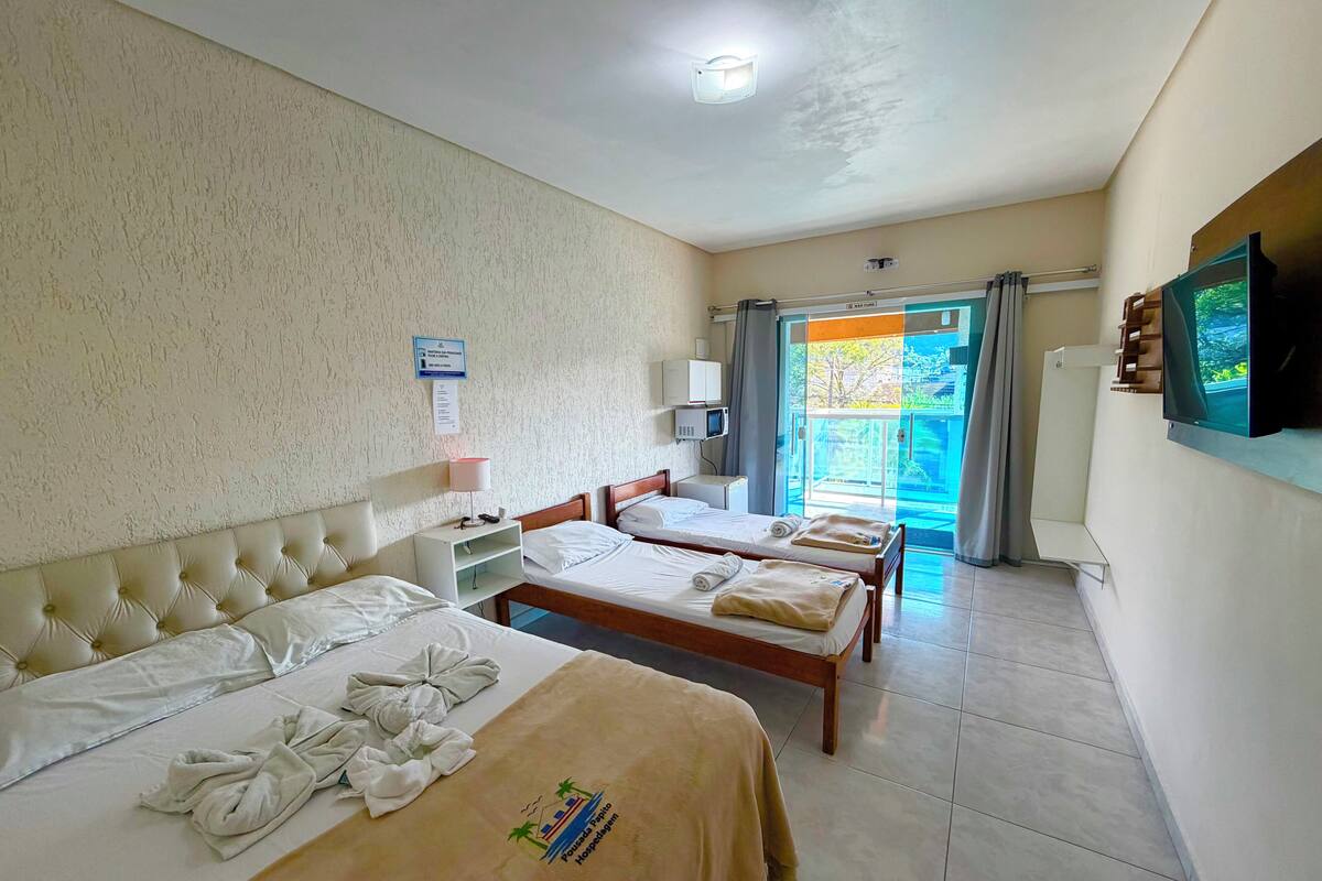 Quarto de hotel com duas camas, televisão, escrivaninha e banheiro visível através de uma porta de vidro.
