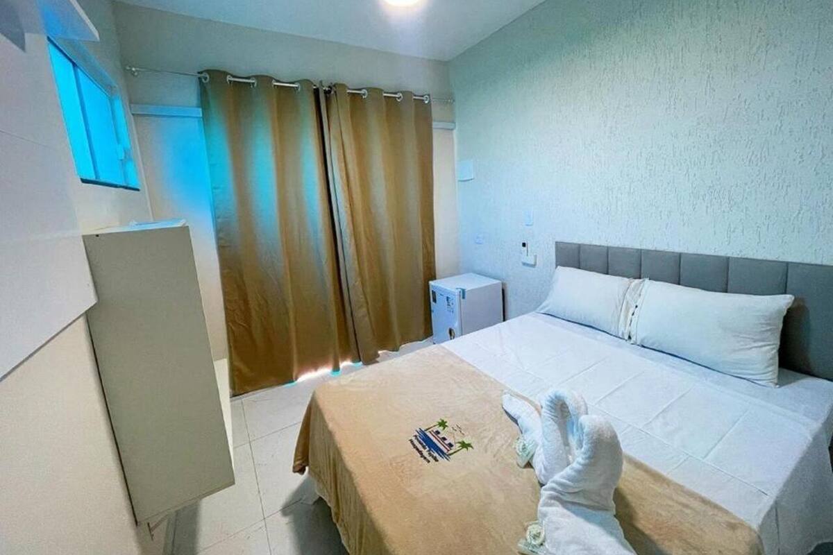 Quarto de hotel com cama, mesa de cabeceira, escrivaninha, cadeira, janela e cortinas.