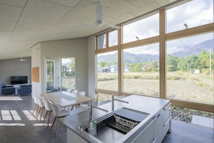 Cozinha moderna com uma ilha grande, área de jantar com mesa e cadeiras brancas e janelas amplas com vista para montanhas e vegetação.