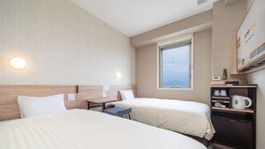 Quarto de hotel com duas camas, uma mesinha, uma criadeira, uma janela e uma estante com amenities.