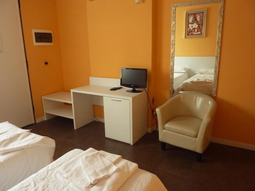Quarto de hotel com uma cama de solteiro, uma escrivaninha com computador, uma cadeira e um espelho na parede.