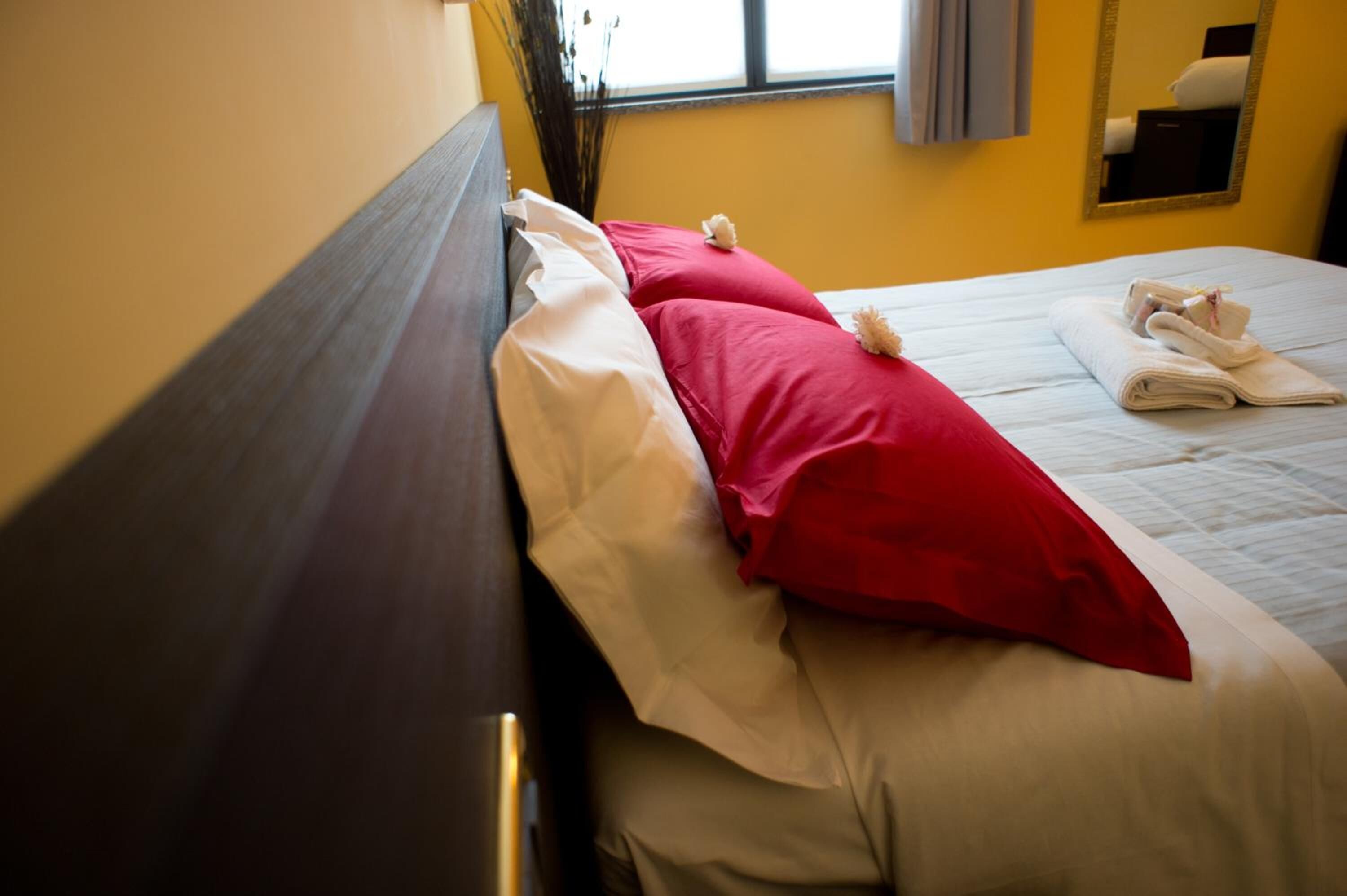 Uma cama bem arrumada com roupa de cama branca e vermelha, cabeceira de madeira e uma janela com cortinas.