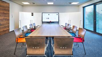 Sala de conferência com uma mesa comprida, cadeiras, tela de projetor e uma janela grande.