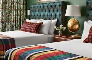 Quarto de hotel com cabeceira estofada, cama branca com manta colorida, mesa de cabeceira com abajur e cortinas florais.