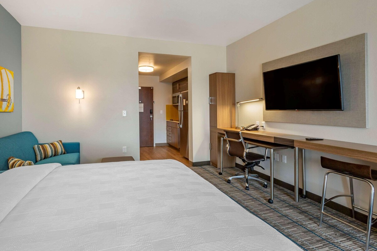 Quarto de hotel com cama, escrivaninha com cadeira, televisão e kitchenette.