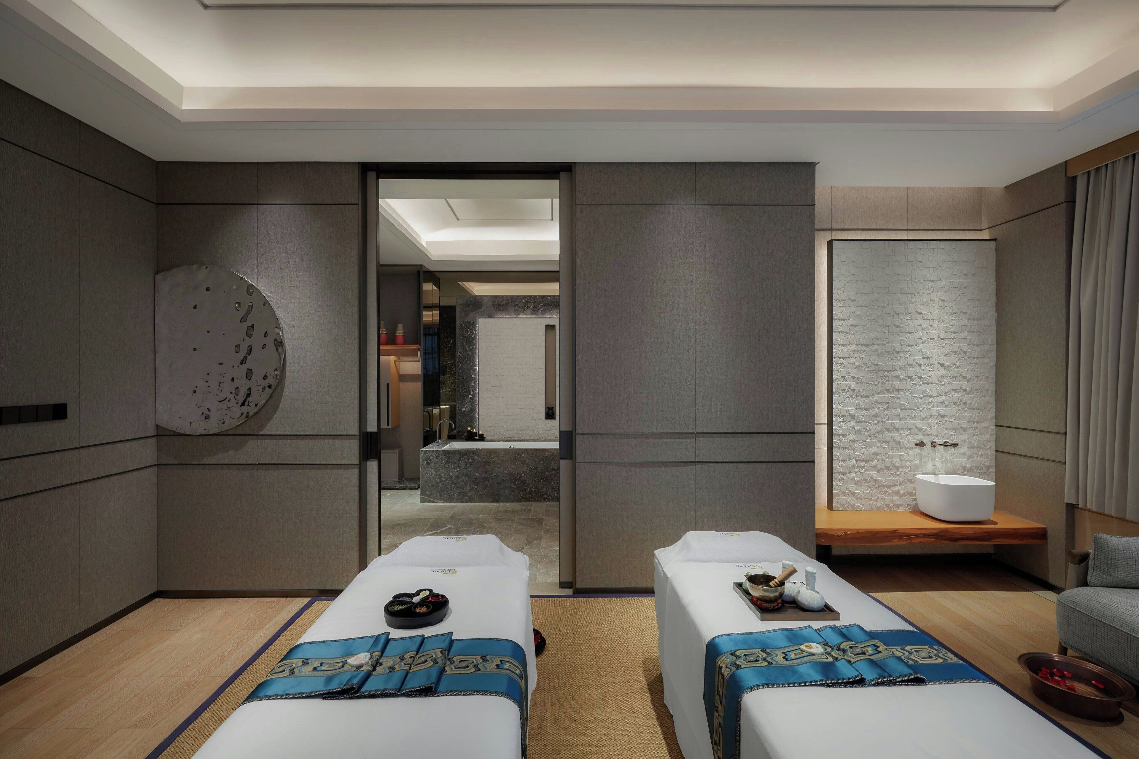 Quarto de hotel moderno com duas macas de massagem, um espelho grande, parede de pedra e pia com cuba branca.