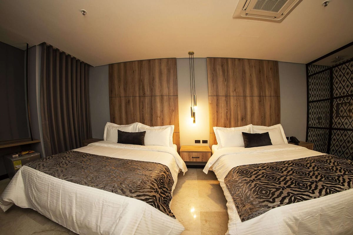 Quarto de hotel com duas camas, cabeceira de madeira, mesa de cabeceira e colcha estampada.