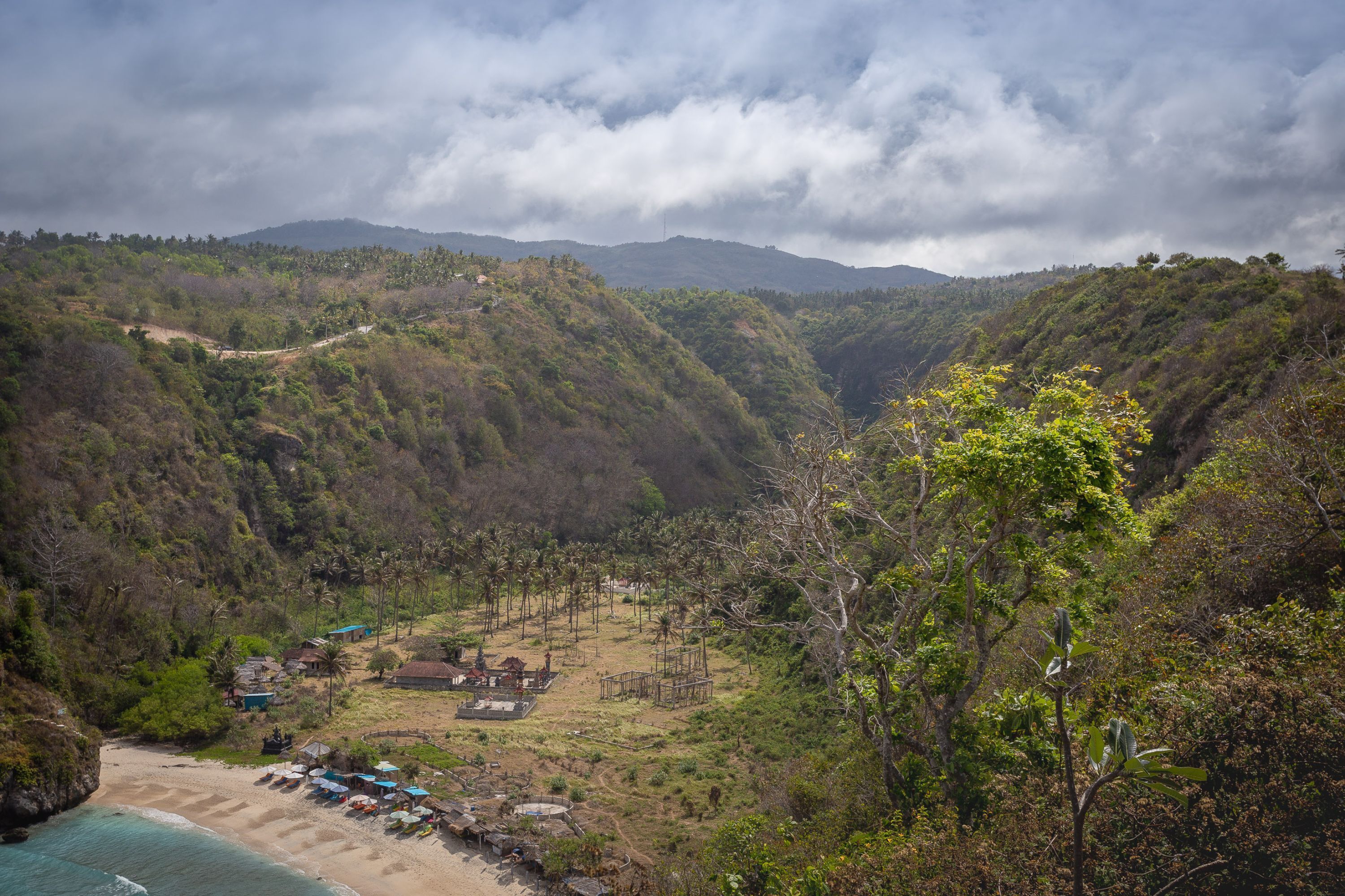 Uma área costeira com praia, um pequeno povoado com edifícios e colinas ao redor.