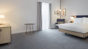 Quarto de hotel com cama, escrivaninha, cadeira, mesinha e janela com cortinas.